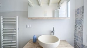 Casa de banho com parede revestida a azulejos brancos, toucador de madeira, lavatório branco em forma de taça grande, frasco azul de champô, espelho e painel de azulejos estampados.