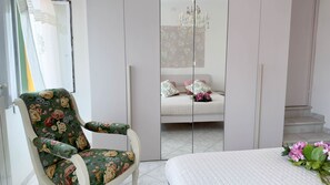 Quarto com poltrona estampada, cama com colcha branca e guarda-roupa espelho.
