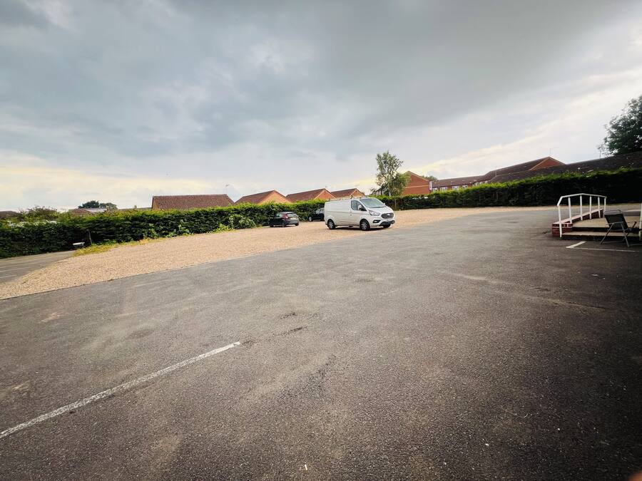 Um estacionamento com uma van branca e um carro preto. Casas ao fundo e um sebe ao lado.