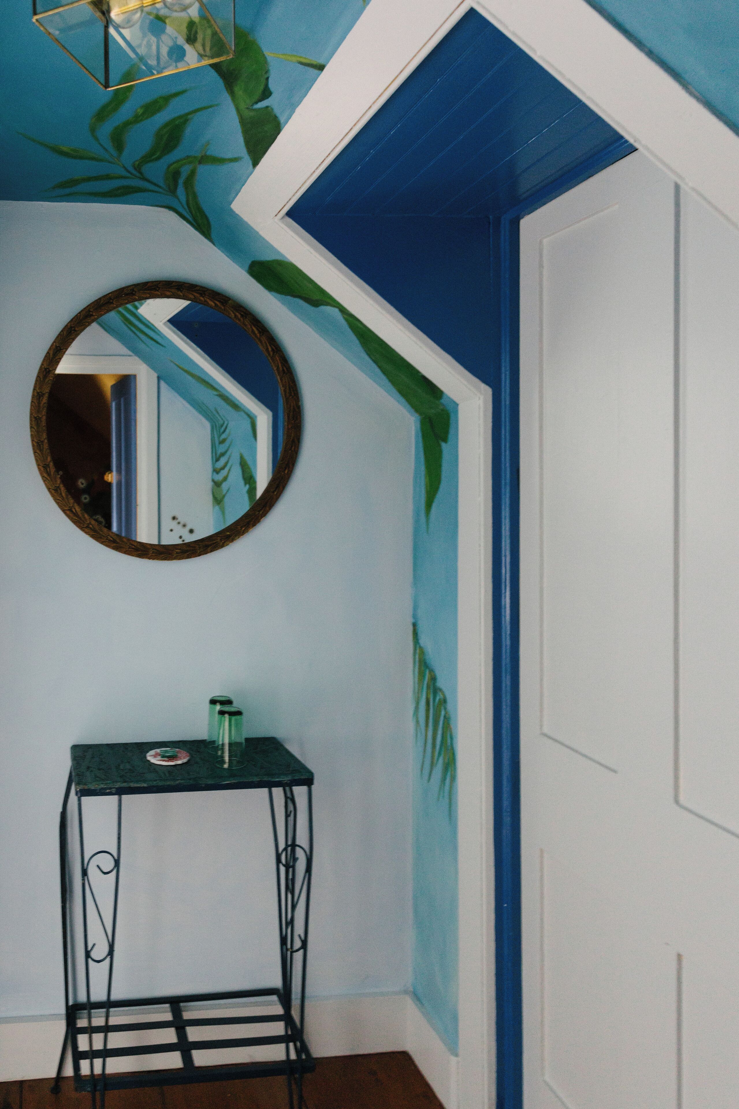 Quarto com esquema de cores azul e branco, espelho redondo na parede e uma pequena mesa de metal preto com uma garrafa sobre ela.