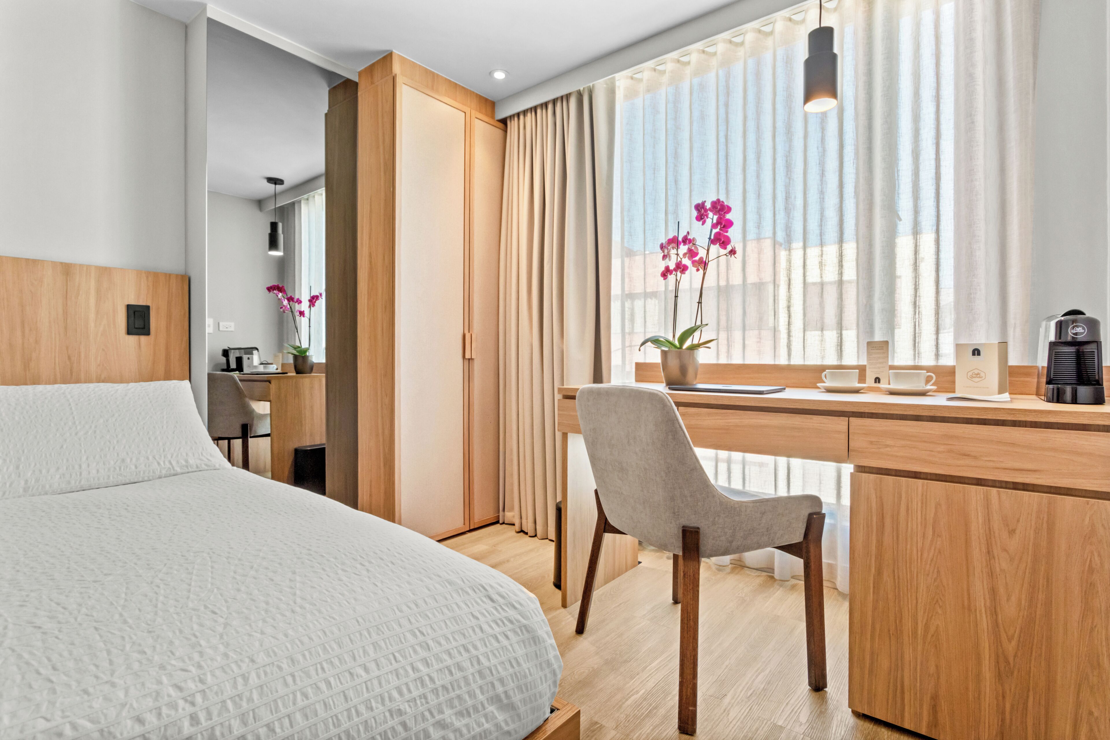 Quarto de hotel com cama, escrivaninha, cadeira, máquina de café e uma planta orquídea.