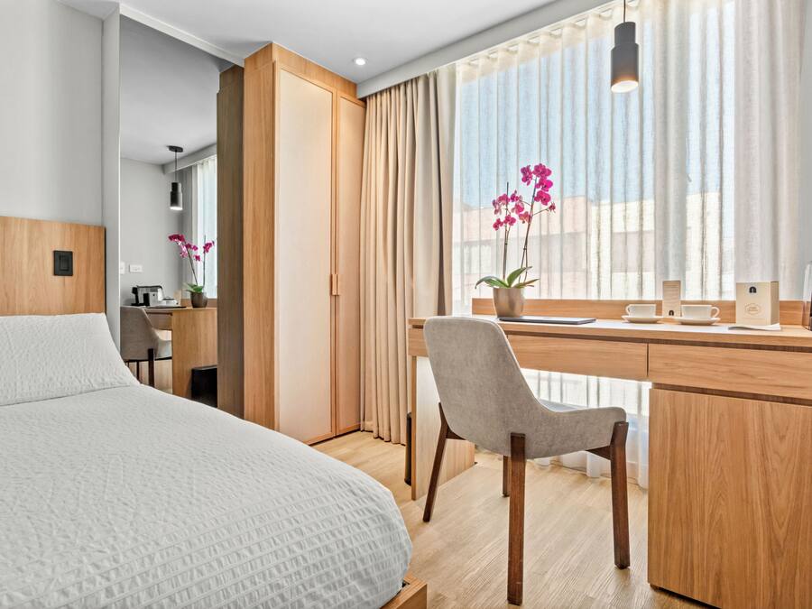 Quarto de hotel com cama, escrivaninha, cadeira, máquina de café e uma planta orquídea.