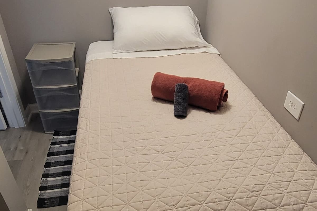 Uma cama de solteiro com colchão acolchoado, uma toalha enrolada e um travesseiro branco. Há um guarda-roupa branco acima da cama e um conjunto de caixas organizadoras cinzas à esquerda.