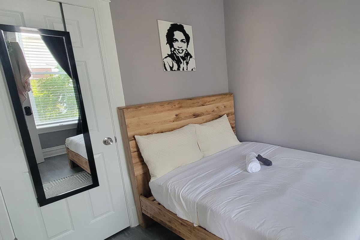 Quarto com cama de madeira, roupa de cama branca, uma pequena mesa de cabeceira e um espelho fixado na parede.