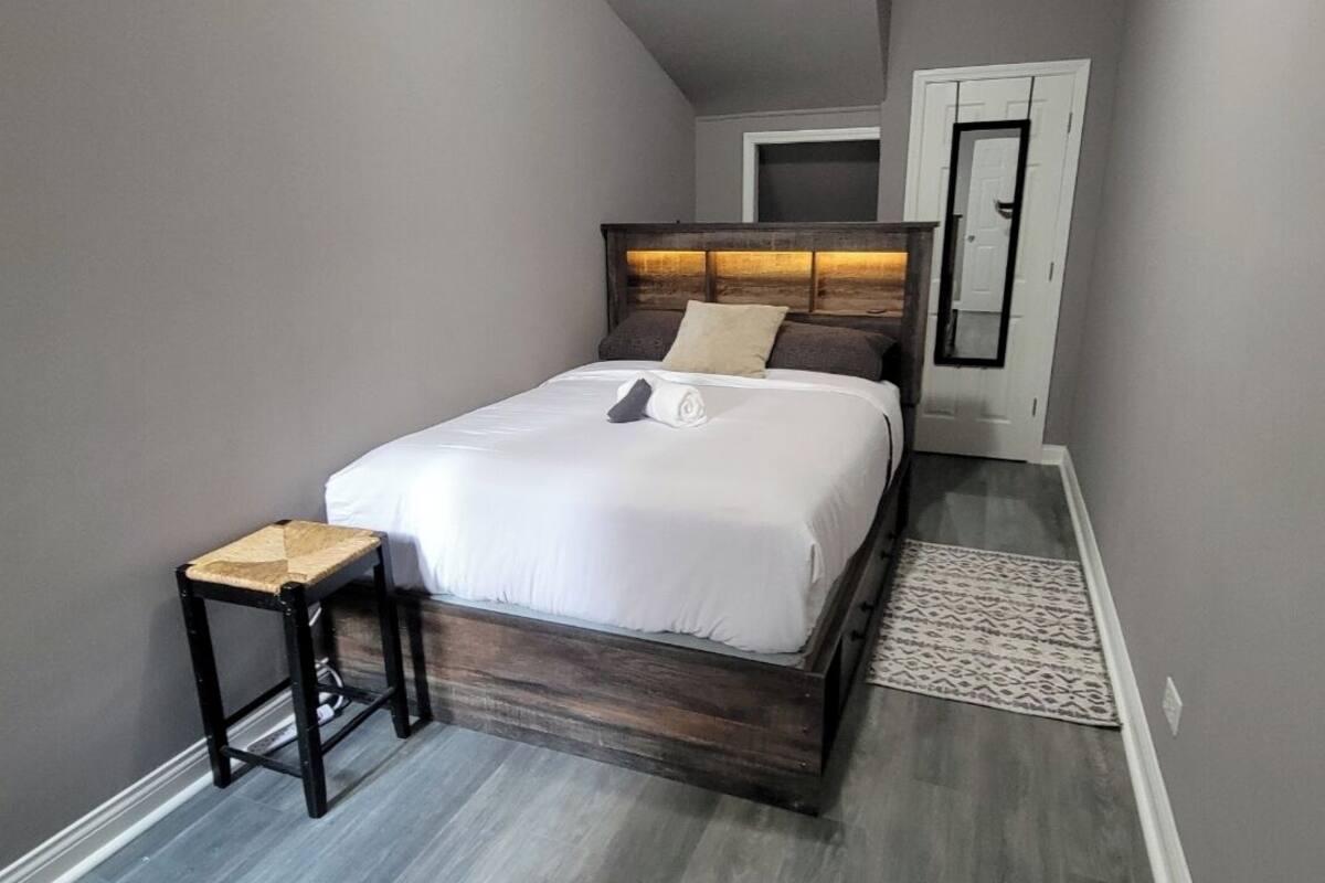 Quarto moderno e estreito, com uma cama, um banquinho pequeno e uma porta.
