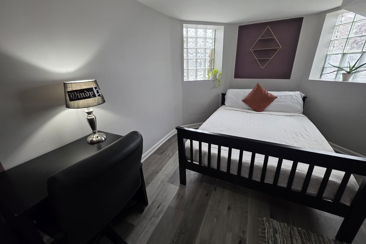 Quarto com cama, escrivaninha e abajur. Janelas com cortinas e um objeto decorativo acima da cama.