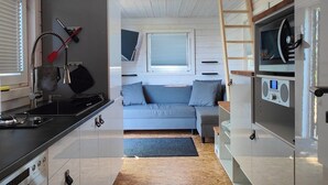 Cozinha compacta e moderna, equipada com fogão embutido, micro-ondas e pia. Uma escada de madeira dá acesso a um espaço de dormir no mezanino, com sofá e janela.