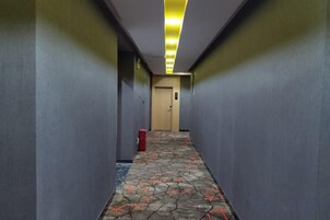 Um corredor com carpete estampado, uma lixeira vermelha e uma porta ao fundo.
