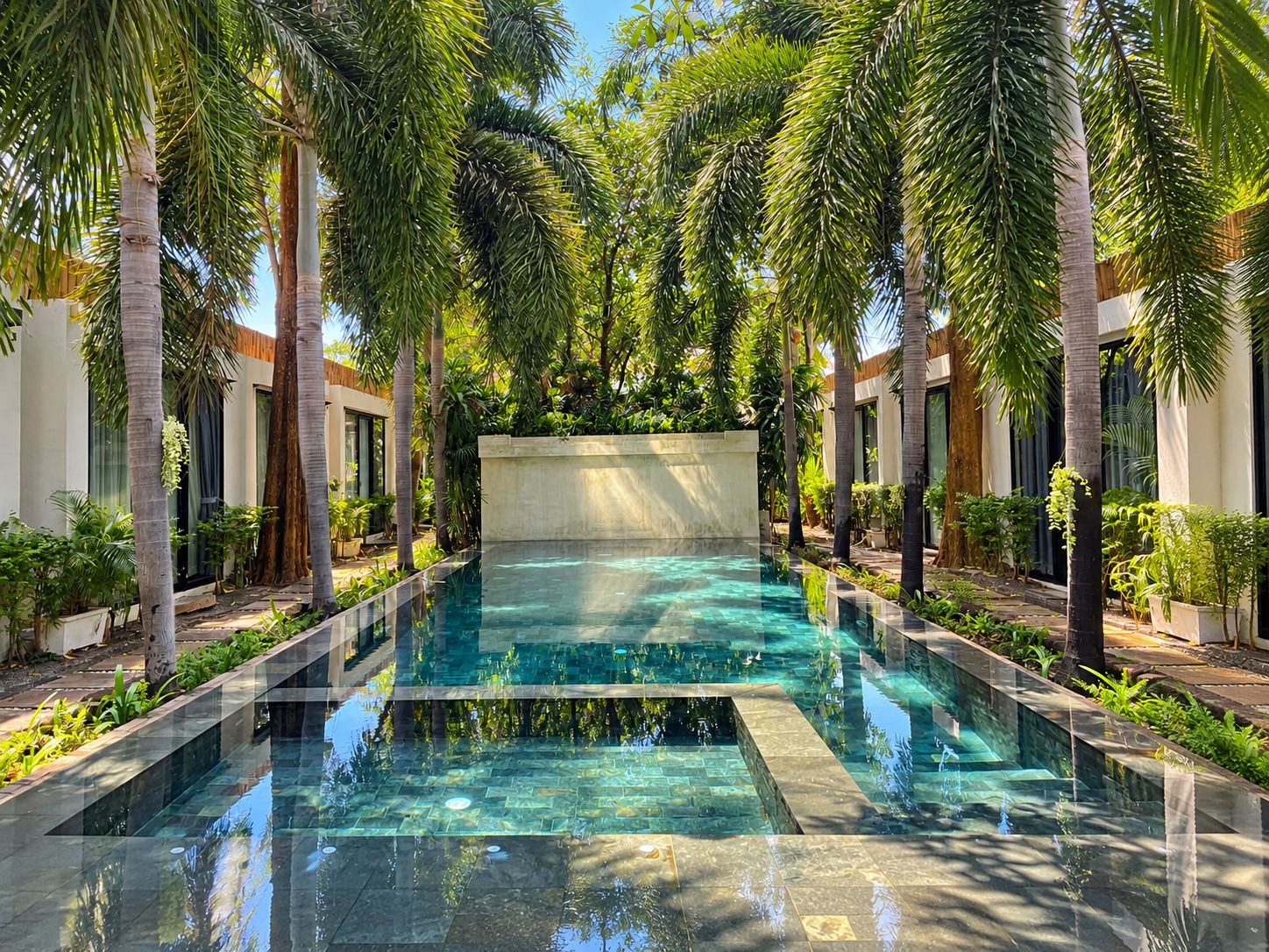 Piscina retangular com água cristalina, cercada por palmeiras e vegetação, com um edifício moderno ao fundo.