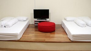 Quarto de hotel com duas camas de solteiro, um banquinho vermelho, uma TV em um suporte e uma parede branca.