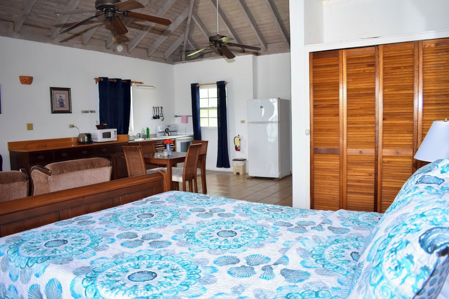 Quarto com cama, guarda-roupa de madeira, área de jantar com cadeiras, mesa e cozinha com frigorífico.
