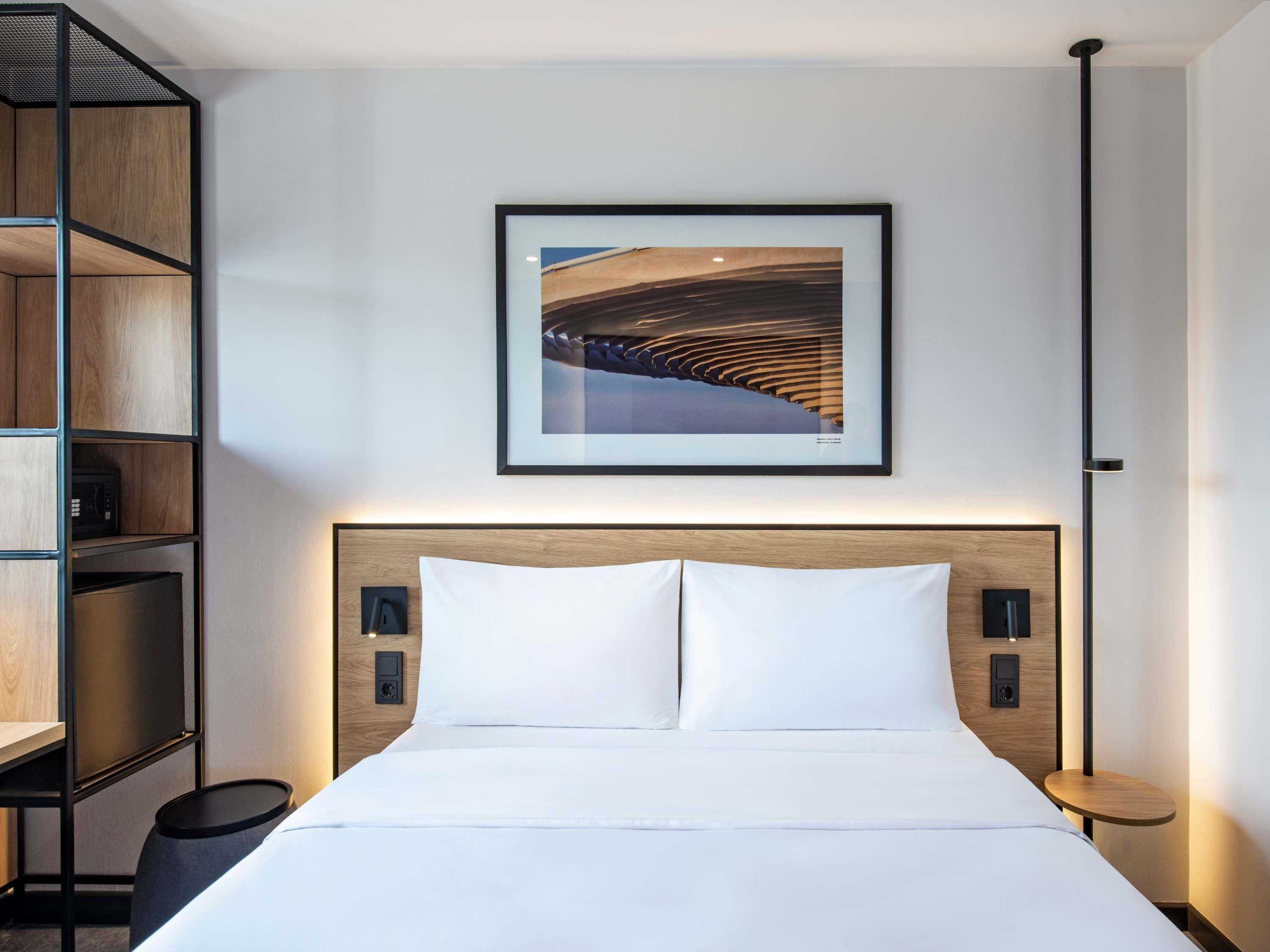 Quarto de hotel moderno com uma cama, um quadro na parede, uma mesa de cabeceira e uma estante de livros.