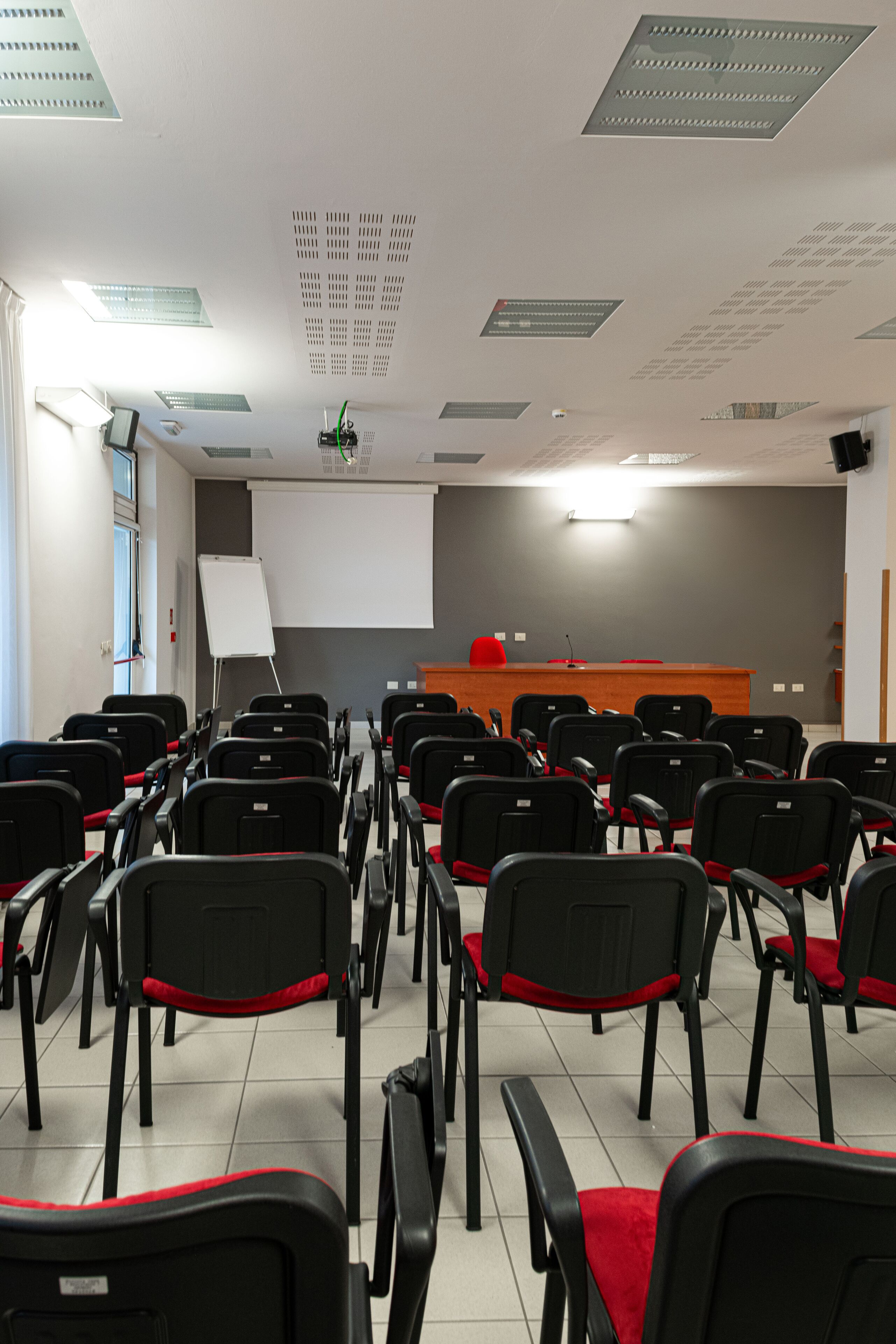 Uma sala de conferências com fileiras de cadeiras pretas com almofadas vermelhas, um quadro branco, uma tela de projetor e um pódio.