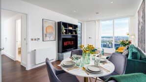 Uma sala de estar moderna com área de jantar, uma janela ampla com vista para a cidade, um rack preto e uma TV de tela plana exibindo 'NETFLIX'.