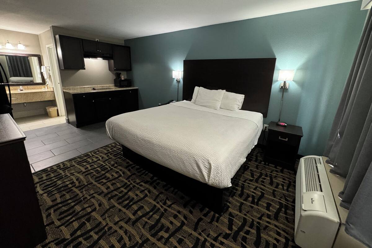 Quarto de hotel com cama, escrivaninha, mesa de cabeceira, luminária e aparelho de ar condicionado.