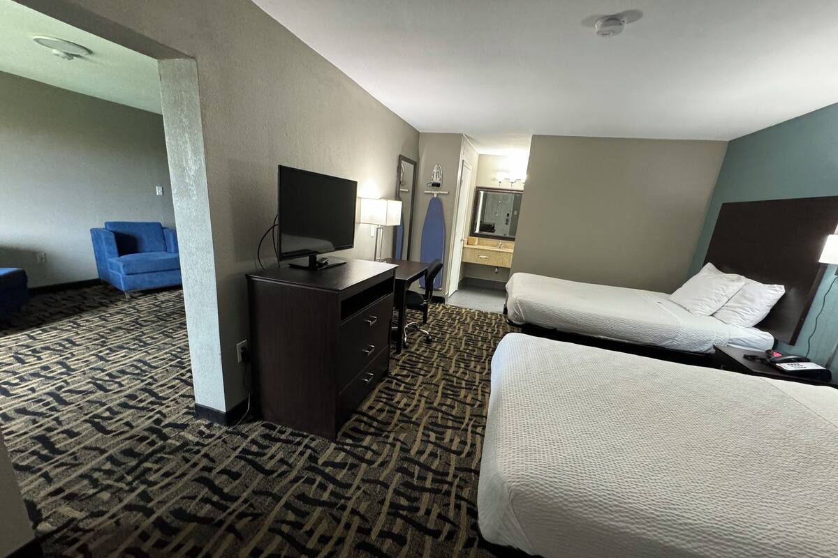 Quarto de hotel com duas camas, uma televisão sobre um aparador, uma escrivaninha com abajur e um banheiro visível através de uma porta aberta.