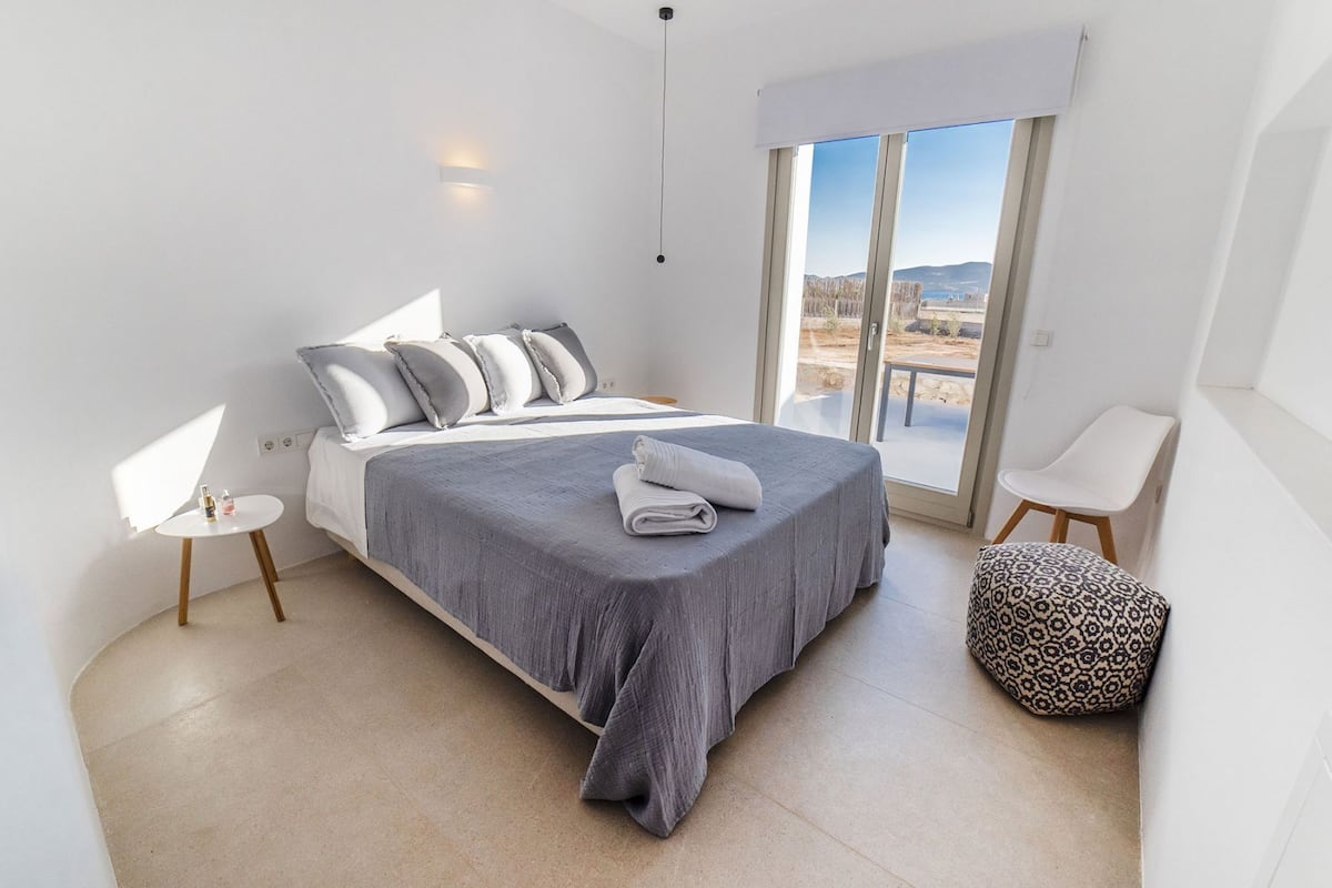 Quarto moderno com uma cama grande, uma mesa pequena, uma cadeira e vista para o exterior.