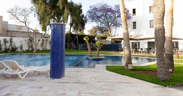Uma área de piscina com espreguiçadeiras, um pilar revestido de azulejos azuis, palmeiras e um edifício ao fundo.