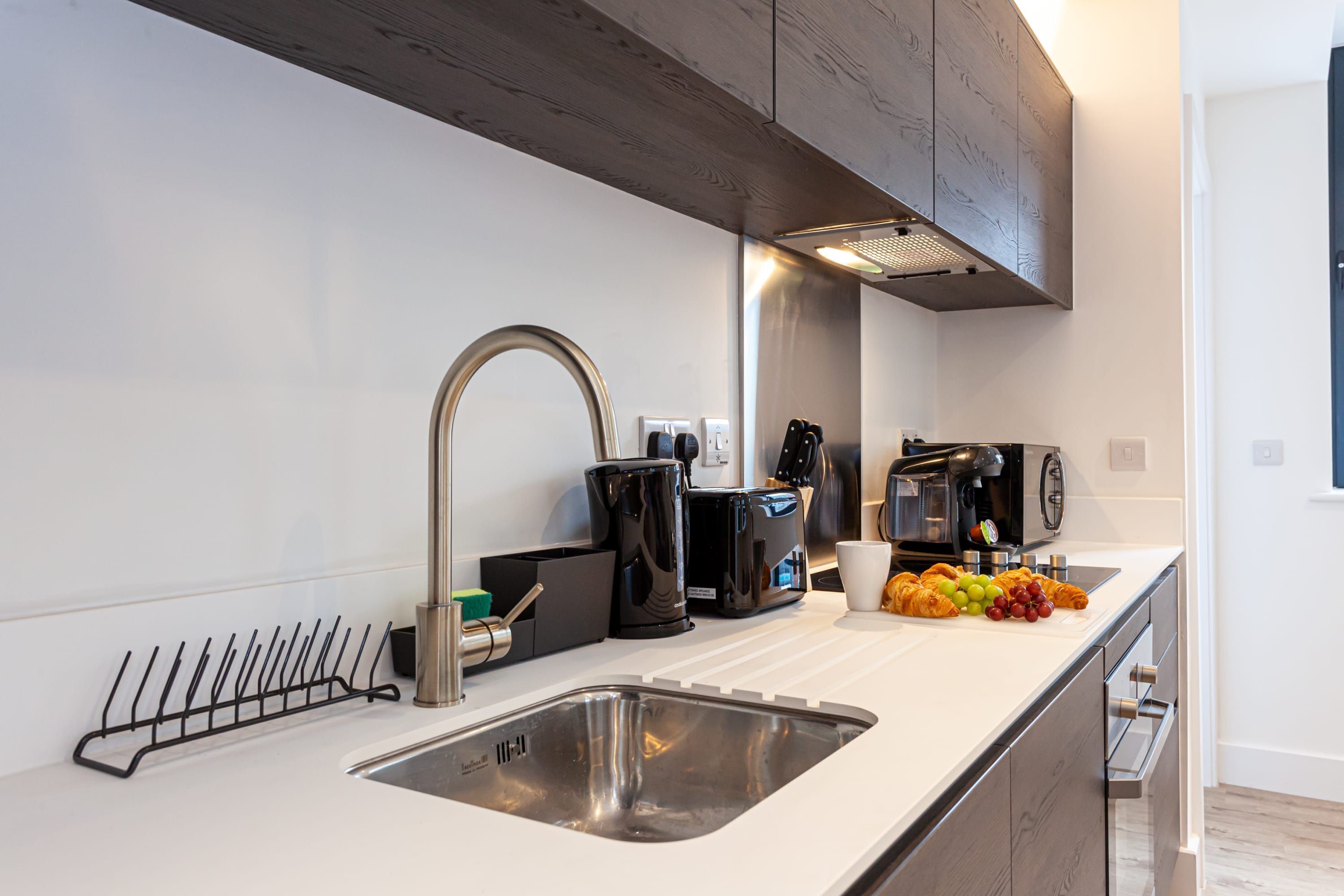 Cozinha moderna com pia de aço inoxidável, eletrodomésticos pretos e bancada branca.