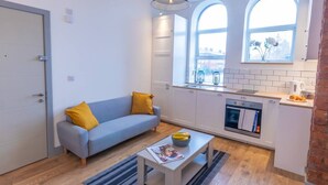 Espaço compacto com sofá, mesa pequena, área de cozinha com forno e janela com vista para a cidade.