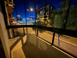 Vista noturna de uma varanda com vista clara da rua, incluindo um prédio com letreiros iluminados e uma construção de vários andares com janelas.