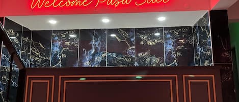 Um balcão de recepção com um letreiro de neon que diz "Welcome Paso Suit" e uma grande parede escura, semelhante a mármore, ao fundo.