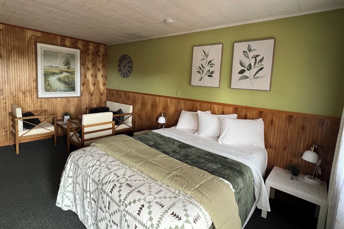 Quarto de hotel com uma cama, duas cadeiras, uma mesinha, uma mesa de cabeceira, um relógio e duas imagens emolduradas na parede.