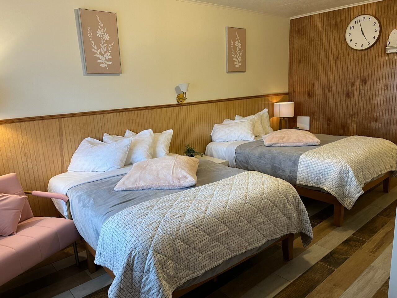 Quarto de hotel com duas camas, uma cabeceira de madeira, um relógio e uma pequena mesa com abajur.