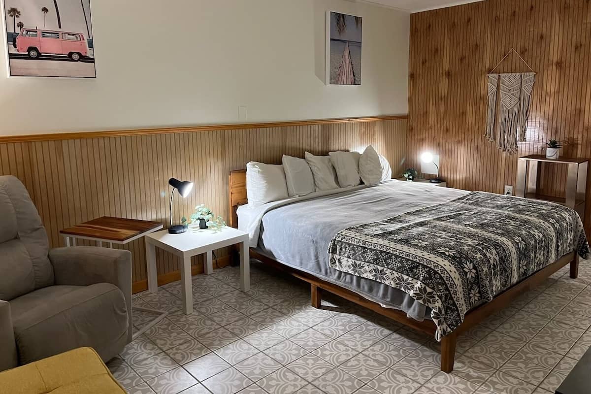 Um quarto com cabeceira de madeira, cama com colcha estampada, mesa de cabeceira com abajur, pequena mesa com uma planta e uma cadeira.