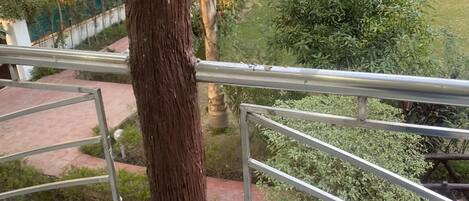 Grade de metal com tronco de árvore em primeiro plano, área pavimentada e vista de um jardim com plantas diversas e um edifício ao fundo.