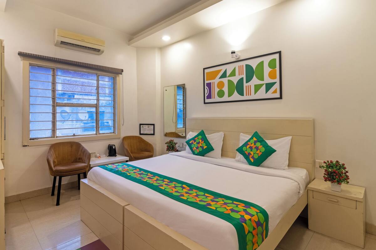 Quarto de hotel com cama, duas cadeiras, mesa de cabeceira, janela com persianas, ar-condicionado e uma obra de arte emoldurada na parede.