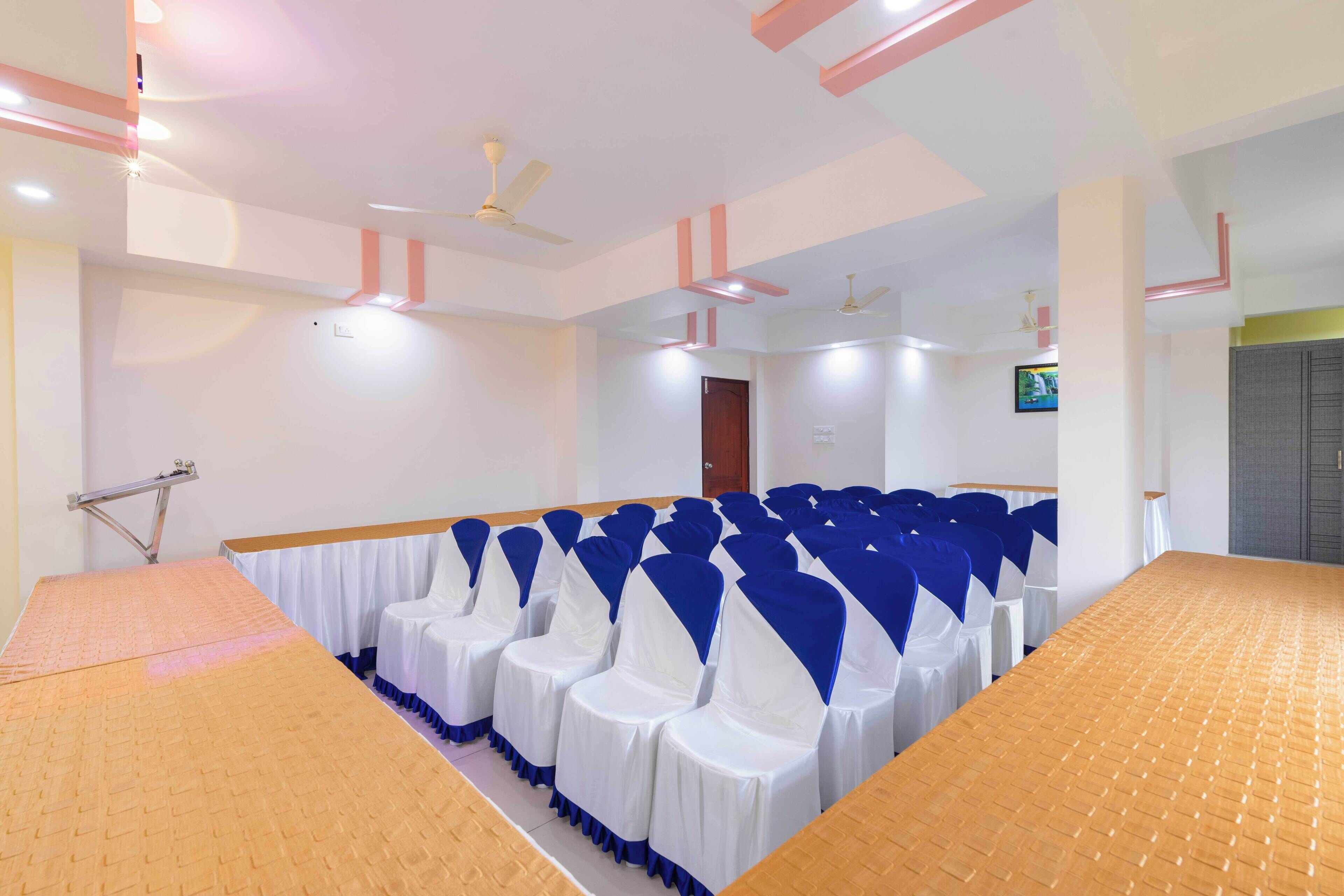 Sala de conferências com fileiras de cadeiras revestidas em branco e azul, uma mesa de madeira, ventiladores de teto e iluminação embutida.