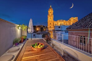 Terraço no telhado com mesa de madeira, plantas em vasos e vista de um edifício histórico com torre e crescente lunar no céu.