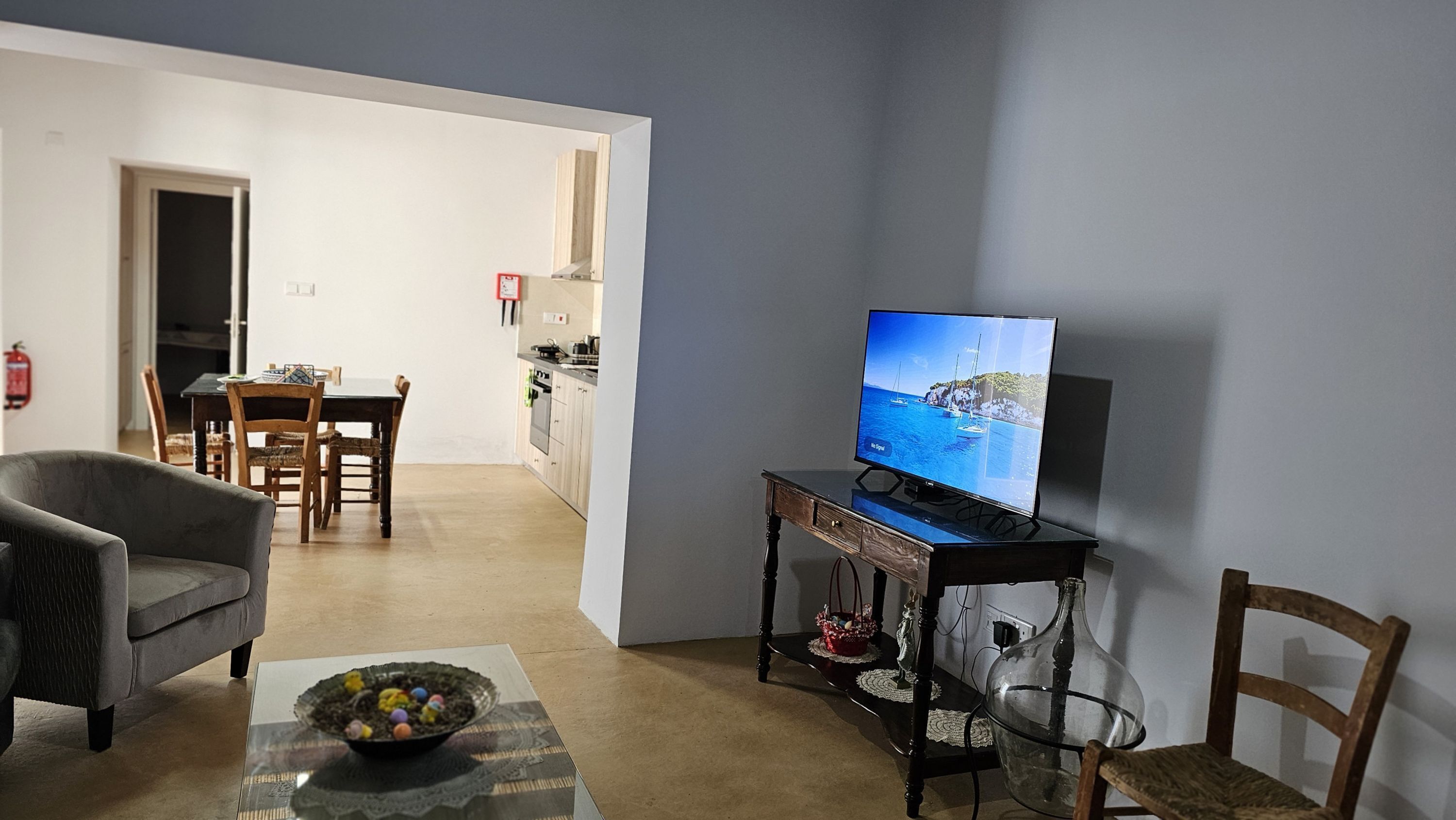 Uma sala de estar com televisão em um suporte de madeira, uma área de jantar com mesa e cadeiras, e uma cozinha ao fundo.