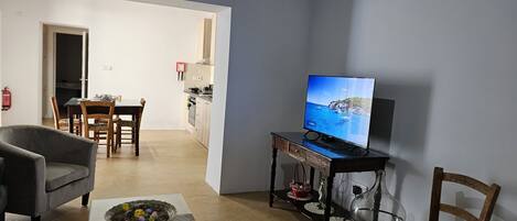 Uma sala de estar com televisão em um suporte de madeira, uma área de jantar com mesa e cadeiras, e uma cozinha ao fundo.