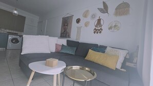 Uma sala de estar com um sofá cinzento, almofadas brancas e estampadas, uma mesa redonda pequena e decorações nas paredes.