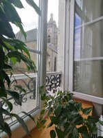 Quarto com uma janela ampla que dá para um edifício histórico, varanda com grades de ferro decorativas e uma planta em vaso no primeiro plano.
