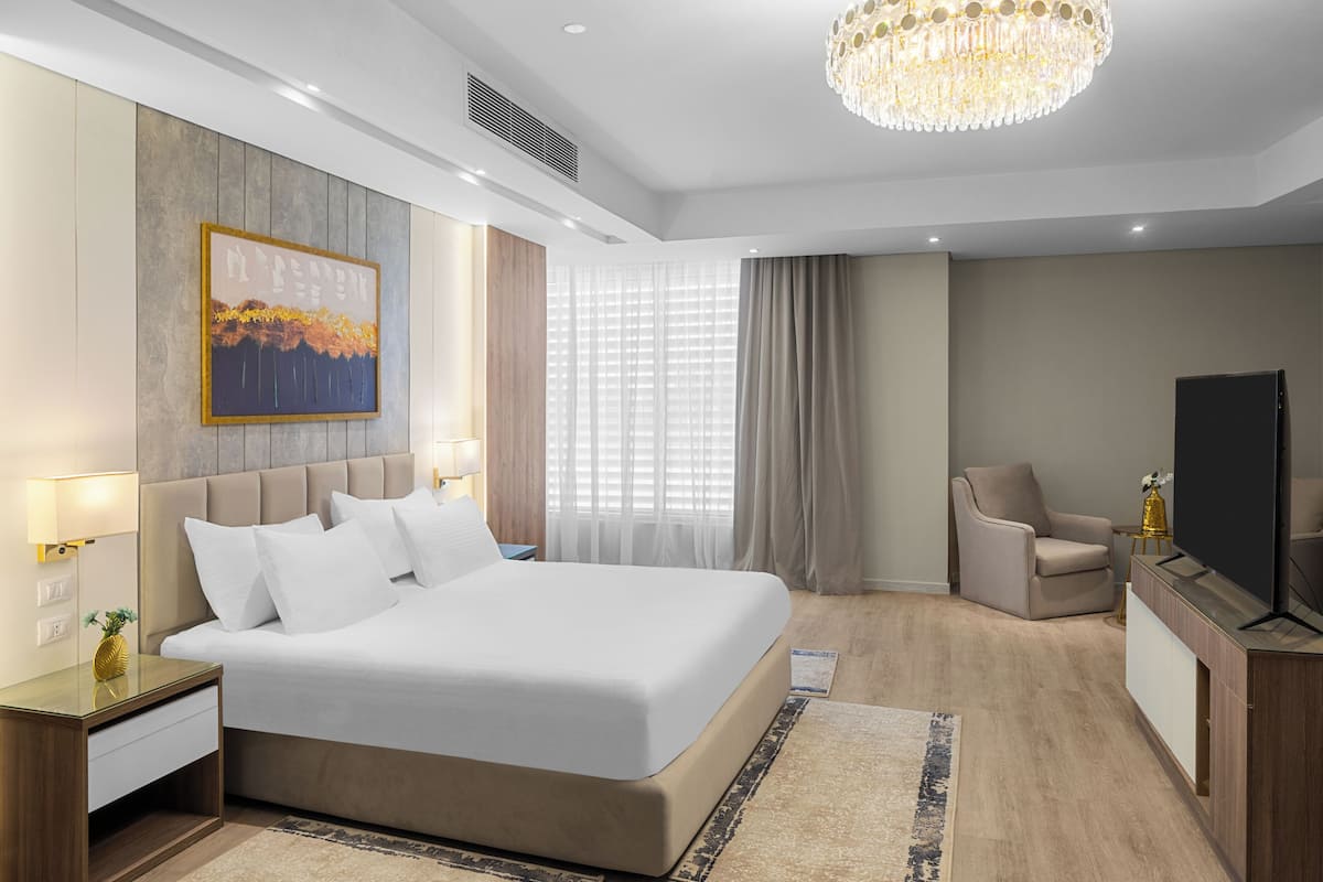 Quarto de hotel moderno com cama grande, escrivaninha, cadeira, televisão e lustre.