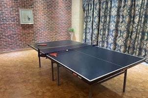 Mesa de pingue-pongue com raquetes, parede de tijolos e cortina estampada.