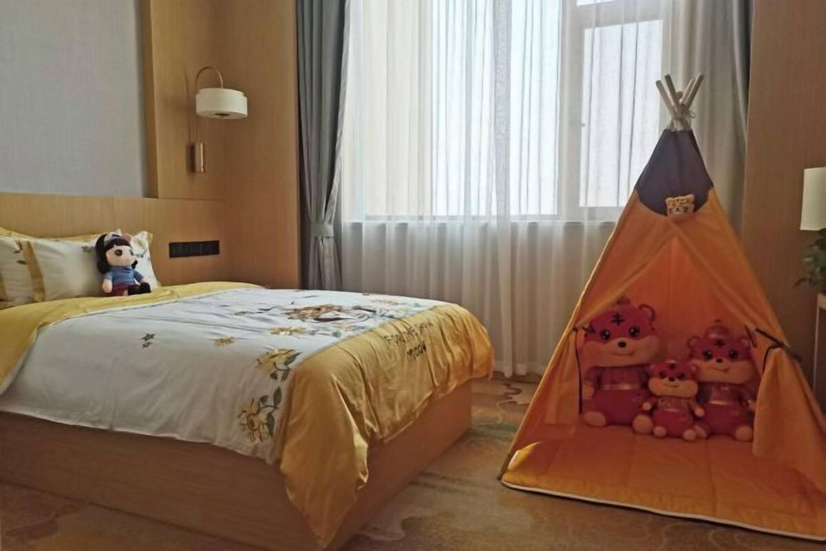 Quarto de hotel com cama, janela com cortinas, luminária fixada na parede e uma tenda tipi com brinquedos macios.