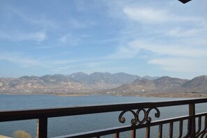 Vista de um lago com montanhas ao fundo, um corrimão de metal em primeiro plano e um céu claro.