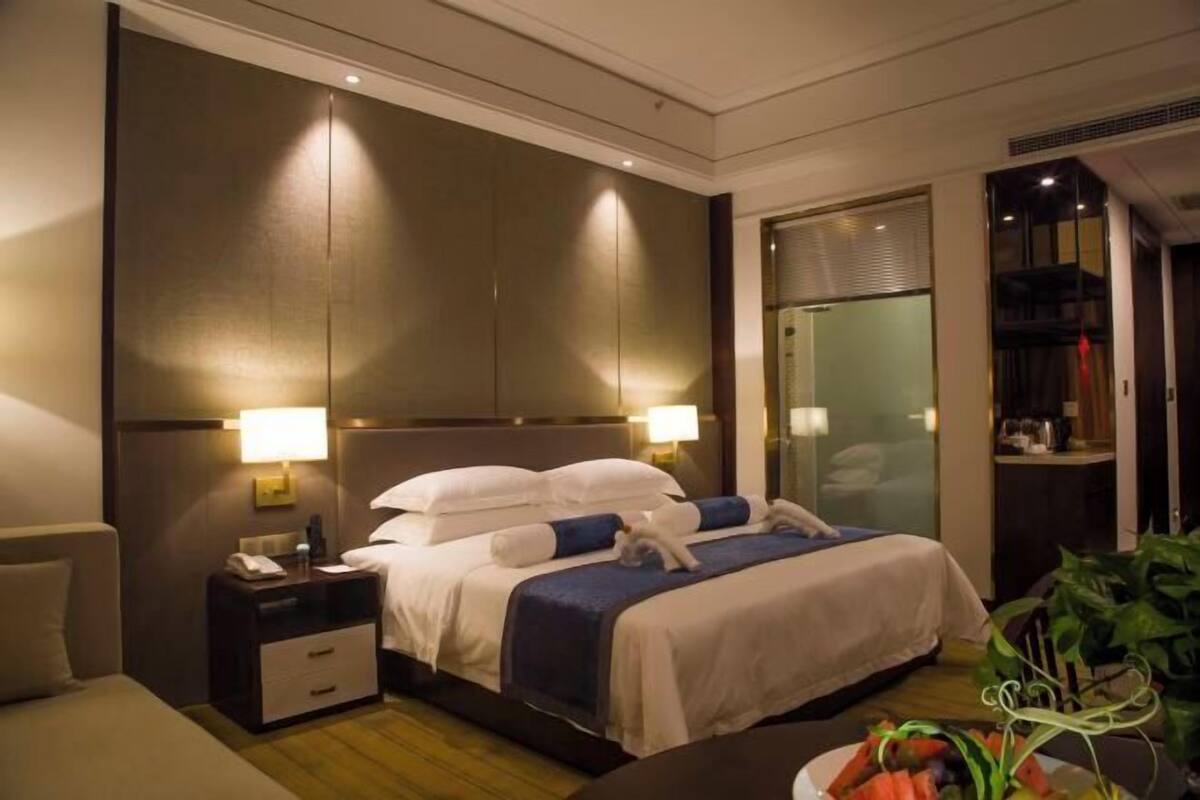 Quarto de hotel com cama, mesas de cabeceira, escrivaninha, cadeira, planta e porta deslizante.