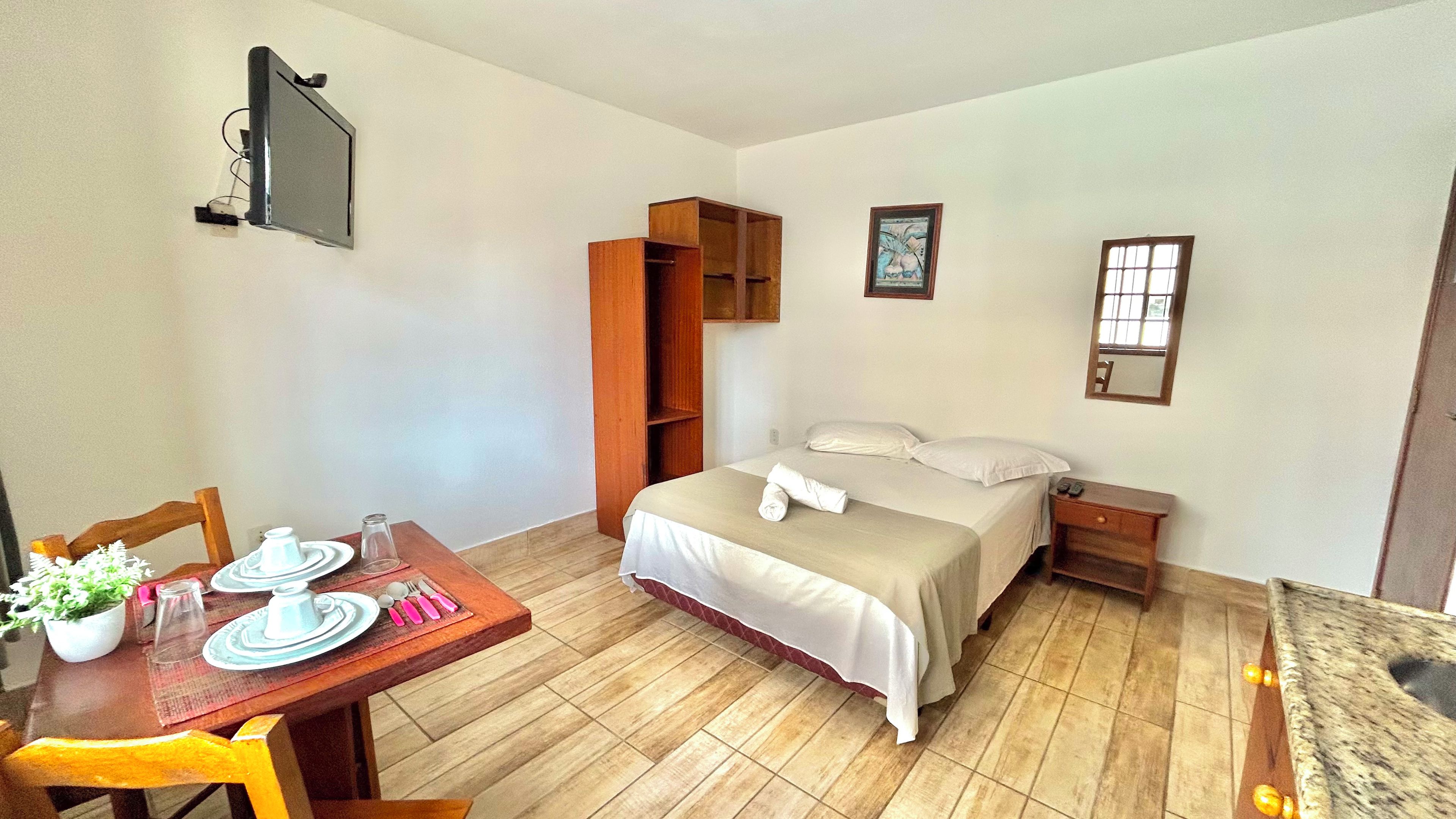 Quarto de hotel com cama, mesa de cabeceira, pequena escrivaninha com cadeira, mesa de jantar com cadeiras e TV fixada na parede.