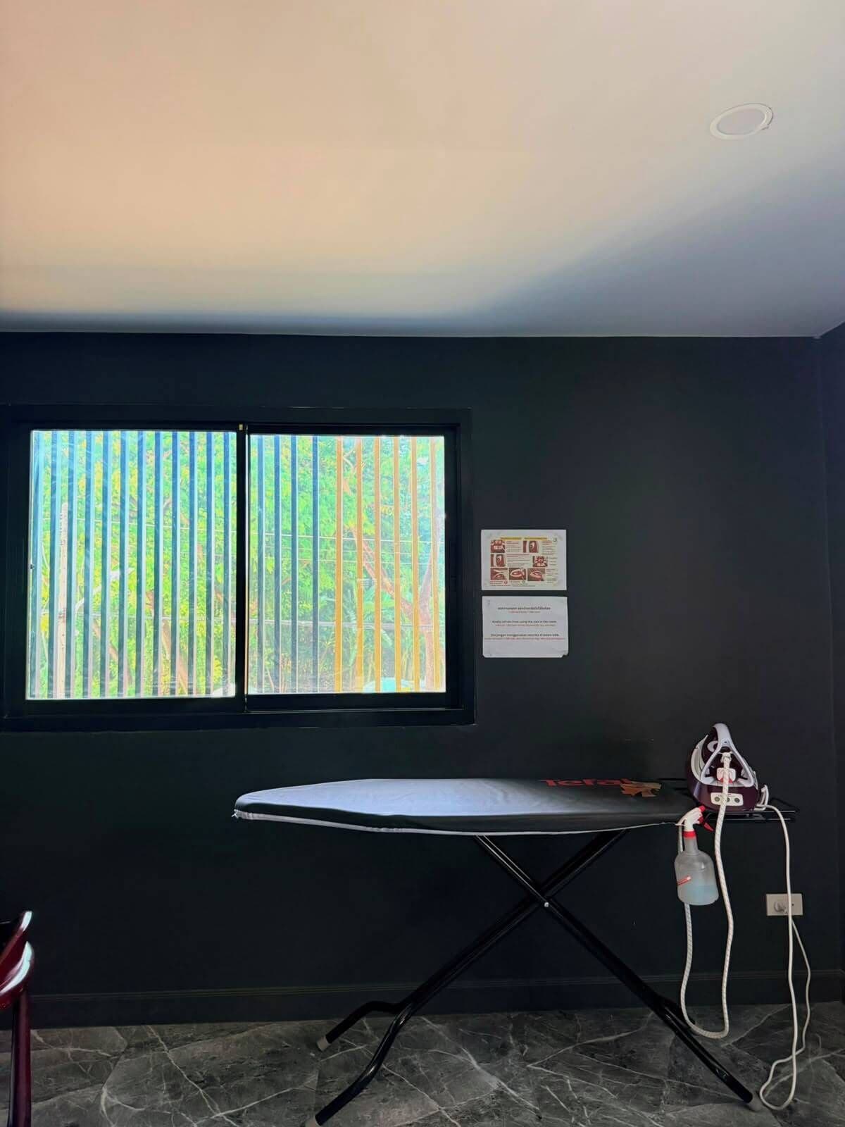 Quarto com uma janela grande, um centro de passar roupa fixado na parede e uma tomada.
