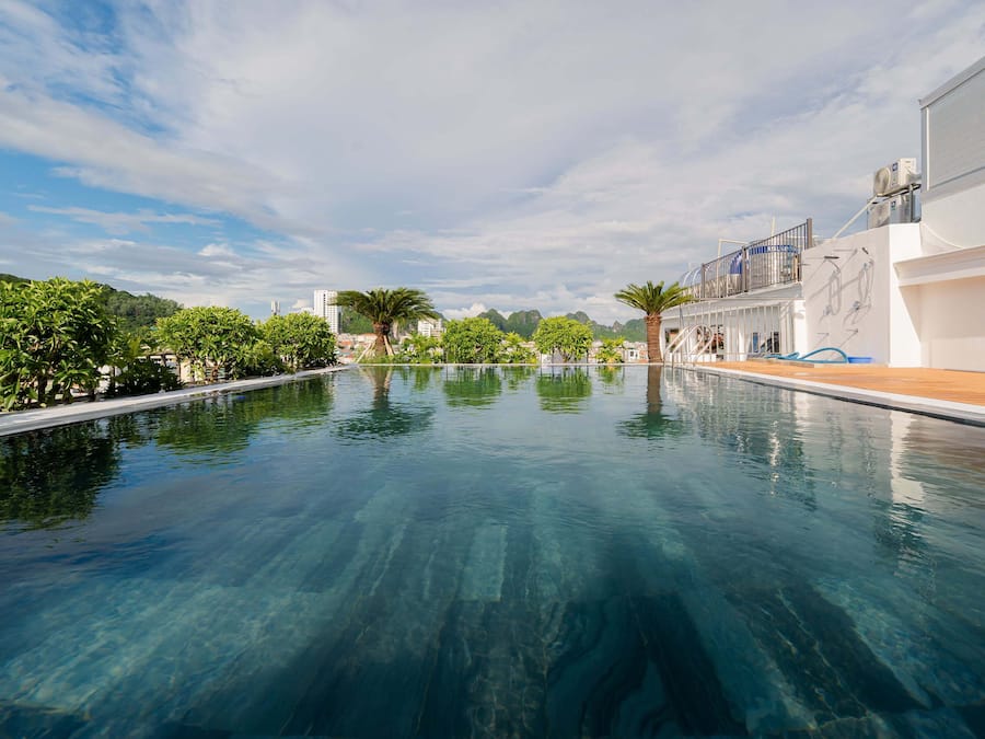 Piscina límpida com deck de madeira de um lado, rodeada por vegetação e edifícios ao fundo.