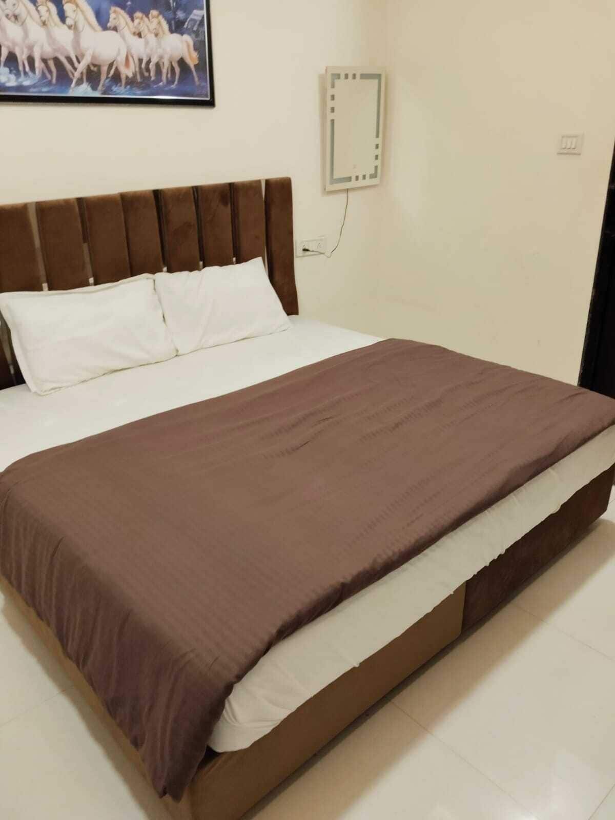 Cama bem arrumada com colcha marrom e almofadas brancas, cabeceira de madeira e luminária fixada na parede.