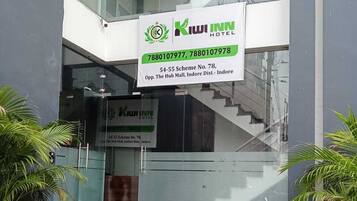 Exterior de um hotel com uma placa exibindo o nome 'Kiwi Inn Hotel' e números de contato. Uma escadaria leva à entrada, e o edifício possui um estilo arquitetônico moderno com janelas de vidro.