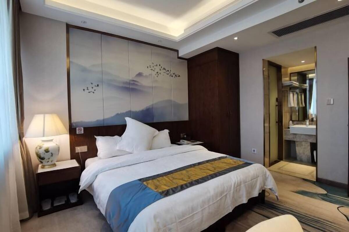 Quarto de hotel com uma cama grande, mesa de cabeceira com abajur, obra de arte na parede e banheiro visível através de uma porta aberta.