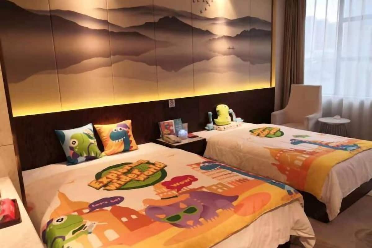 Quarto de hotel com duas camas, cada uma com colchas coloridas com personagens de desenhos animados. Há um grande mural na parede acima das camas, uma pequena mesa com luminária e uma cadeira perto da janela.