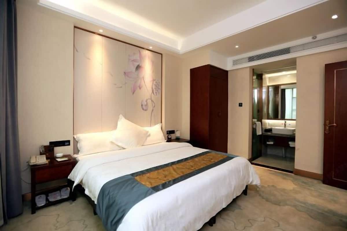Quarto de hotel com uma cama grande, mesa de cabeceira, espelho e banheiro visível por uma porta aberta.